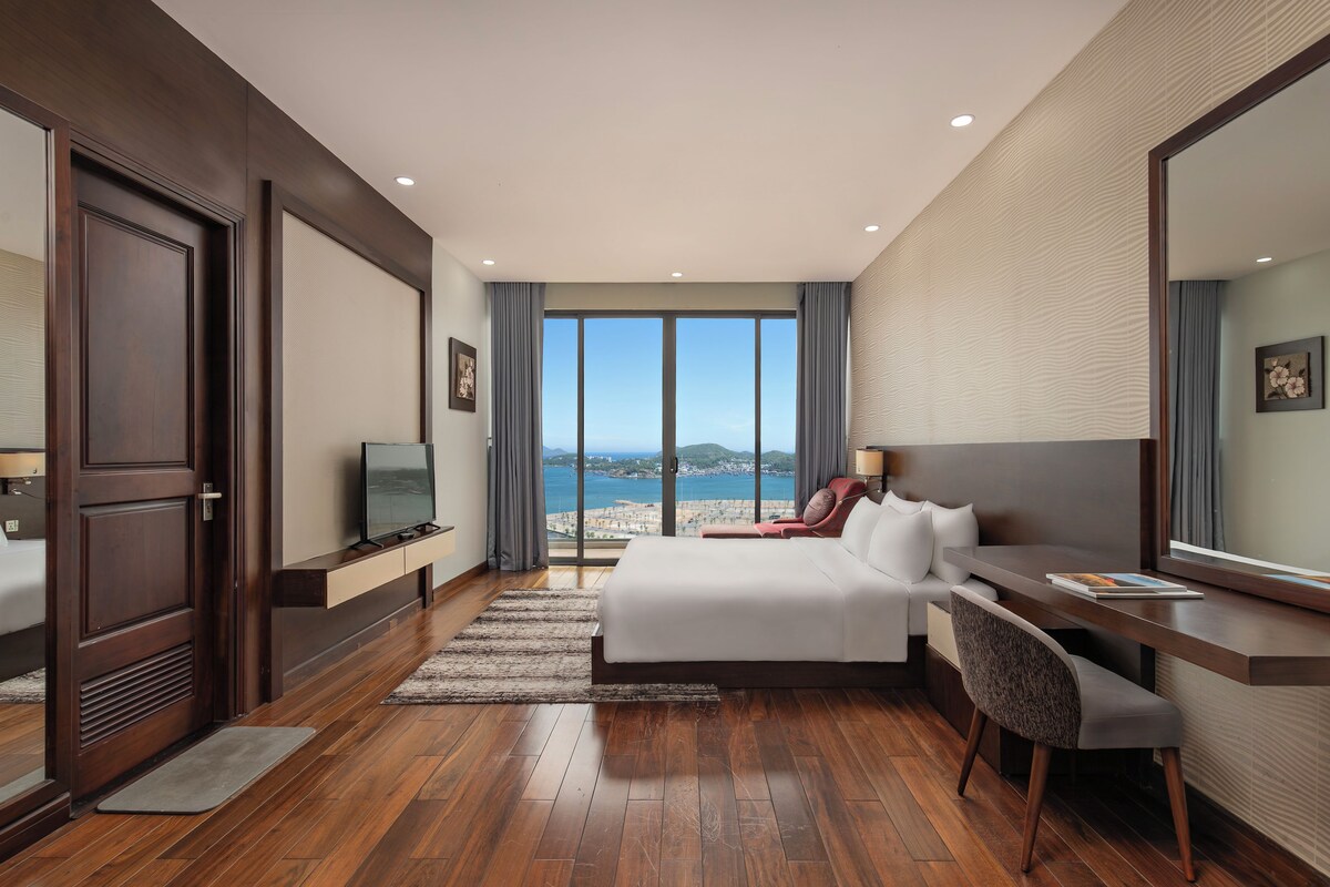 Quarto de hotel com uma cama grande, uma escrivaninha, uma cadeira, uma televisão e vista para o mar.