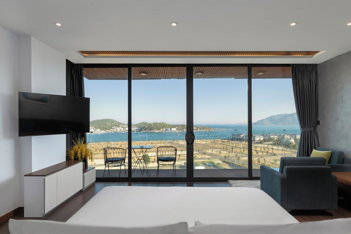 Quarto moderno com cama grande, TV na parede e vista para o mar e para a cidade.