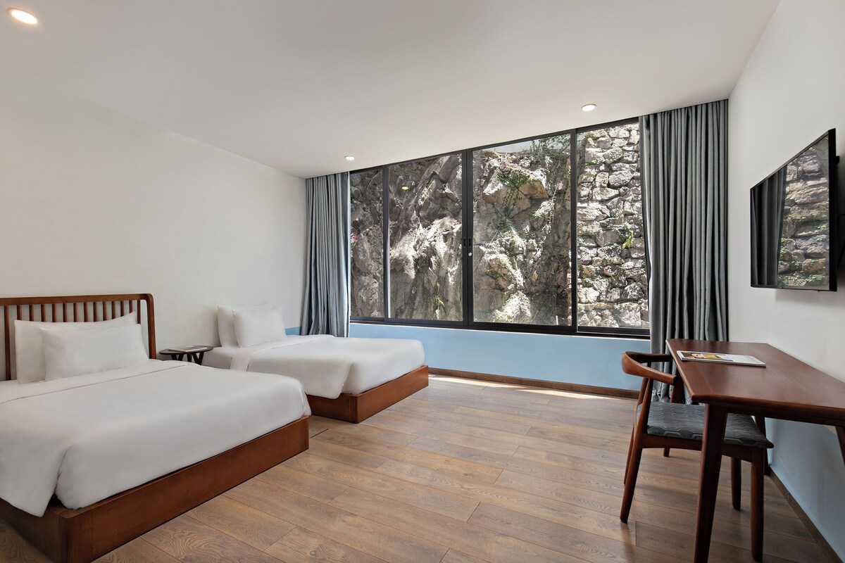 Quarto de hotel com duas camas, uma escrivaninha e uma janela grande com cortinas.