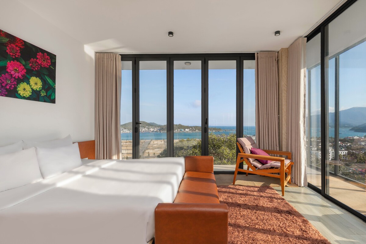 Quarto de hotel moderno com uma cama grande, área de estar e varanda com vista panorâmica.