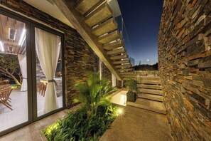 Uma área externa moderna com um muro de pedra, uma porta de vidro que dá para um espaço interno e uma escadaria.