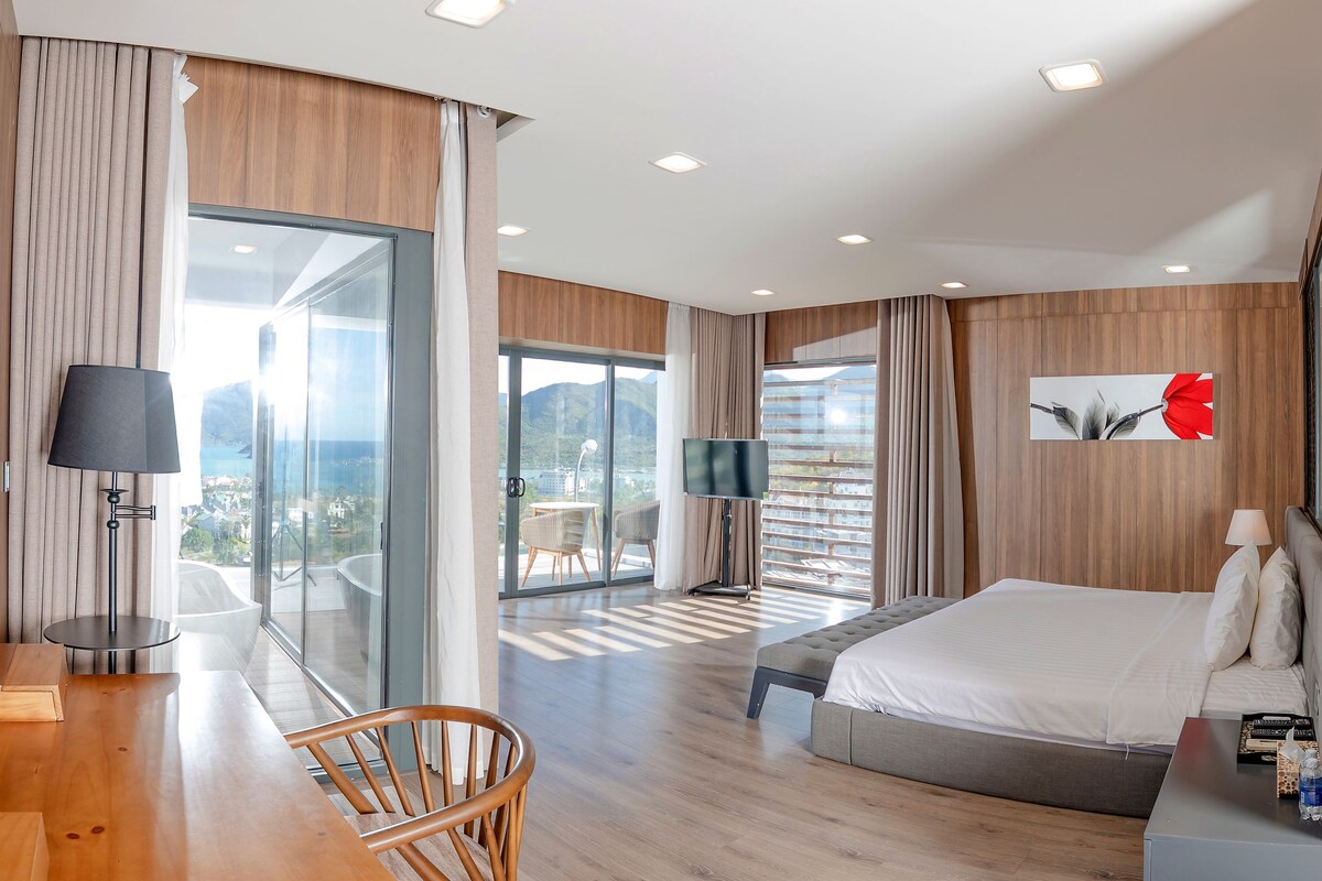 Quarto de hotel moderno com cama grande, escrivaninha, cadeira, televisão e banheiro com chuveiro.