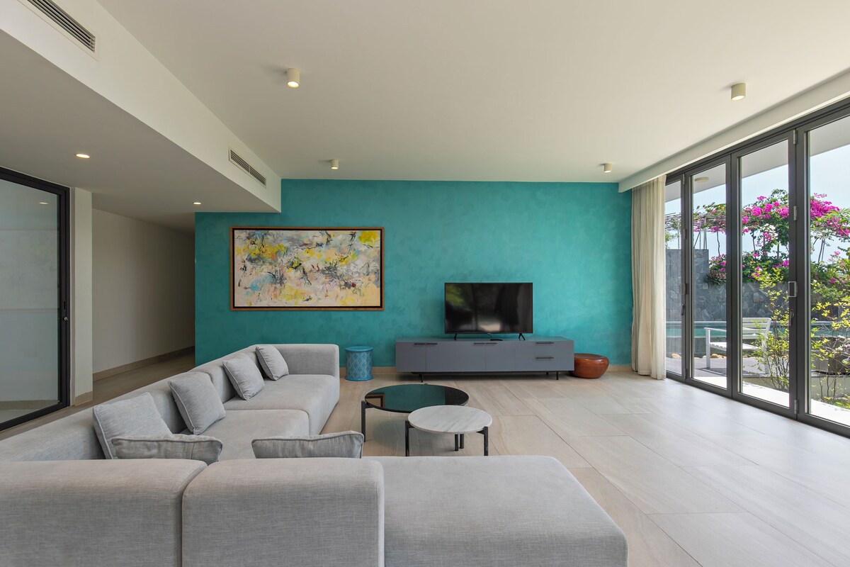 Uma sala de estar moderna com uma parede de destaque em tons de verde-água, um sofá grande, uma televisão e uma vista pela janela.