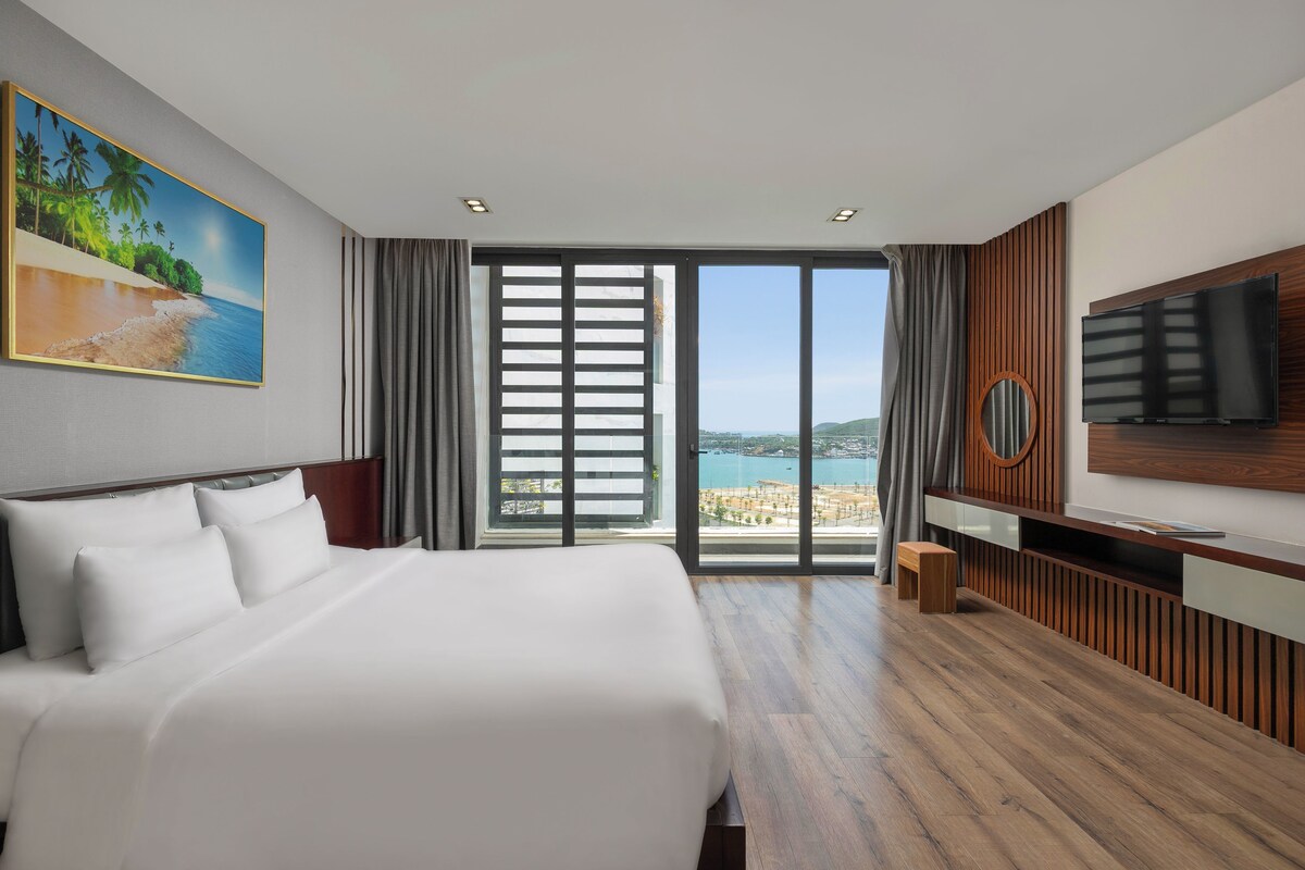 Quarto de hotel com cama grande, televisão, console de madeira e vista para o mar.