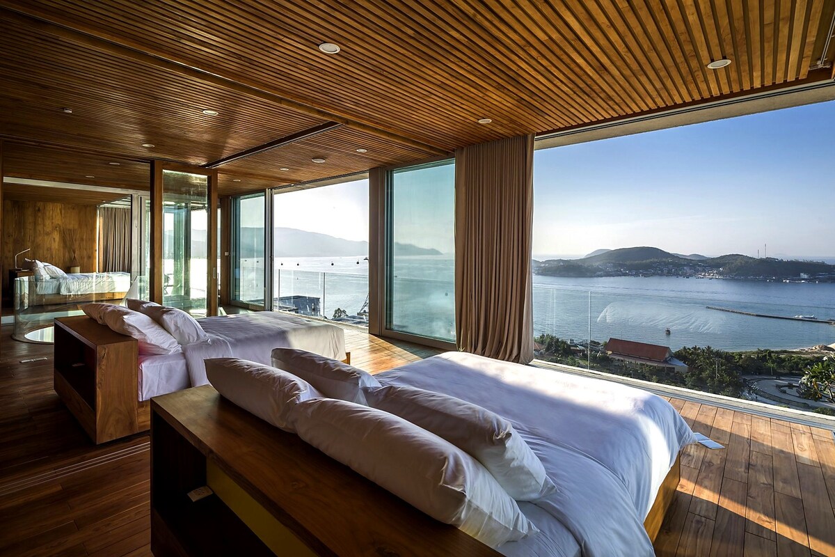 Quarto de hotel moderno com uma cama grande, piso de madeira e vista para o mar.