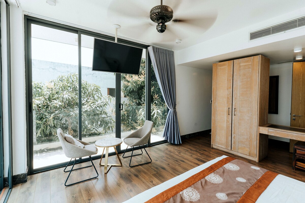 Quarto de hotel moderno com uma grande porta de vidro deslizante, uma TV de tela plana, duas poltronas, um guarda-roupa de madeira e um tapete estampado.