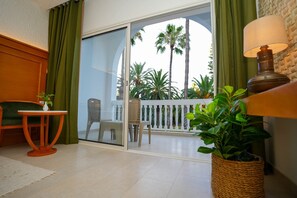 Um quarto com porta de vidro deslizante que dá para uma varanda com guarda-corrimão branco, uma cadeira de vime, uma mesinha com uma planta e um abajur.