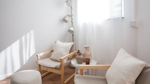 Quarto minimalista com piso de madeira, uma cadeira de madeira, um sofá, uma pequena mesa com um vaso e uma vela, e uma janela com cortinas leves.