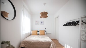 Um quarto com uma cama, duas almofadas, um candeeiro pendente, um porta-roupas e uma parede com duas molduras.