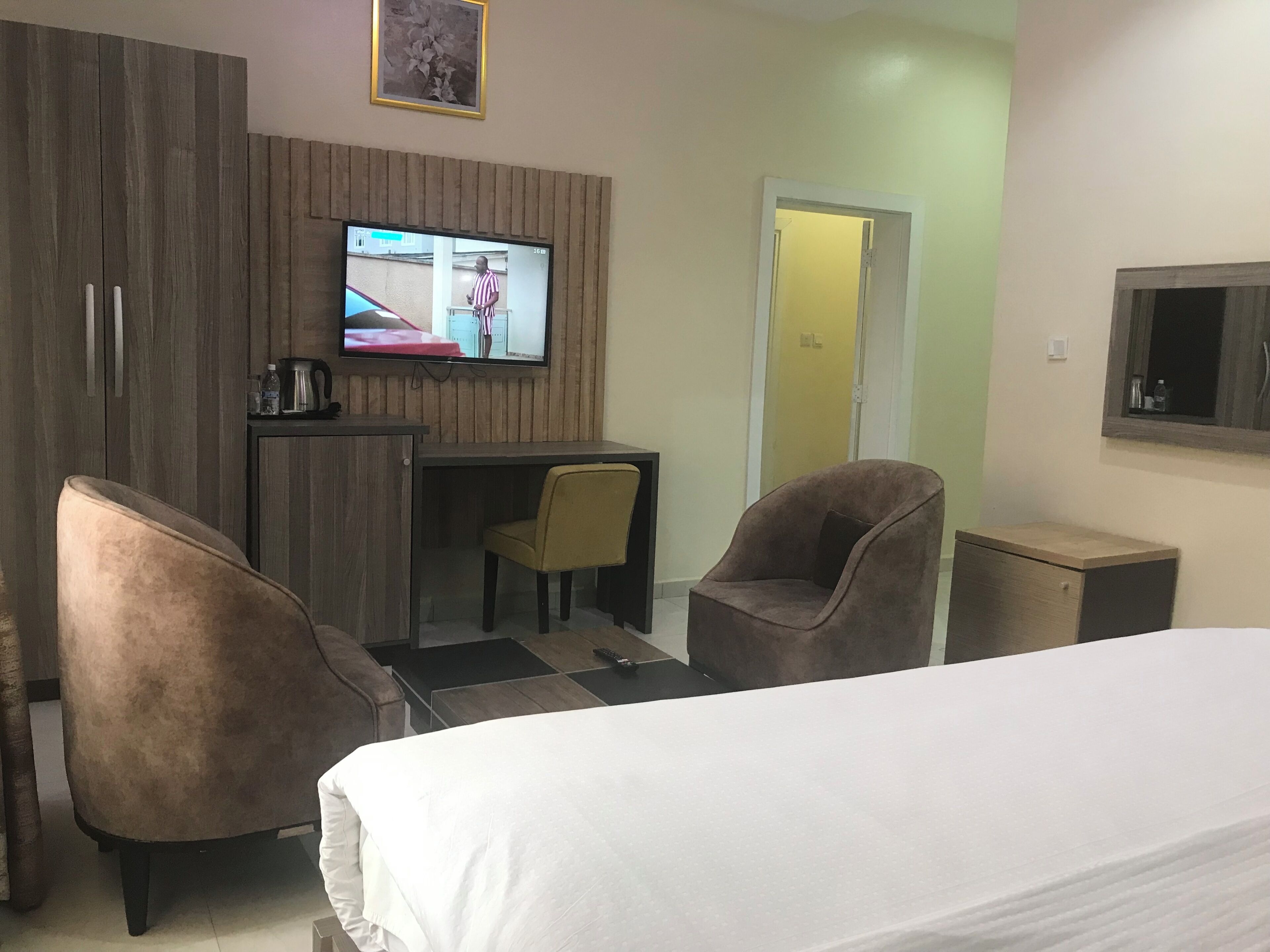 Quarto de hotel com uma cama, duas poltronas, uma secretária com um computador, uma televisão na parede e uma porta que dá para outro espaço.