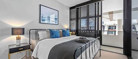 Quarto moderno com cama, mesa de cabeceira, abajur e uma janela grande com persianas.
