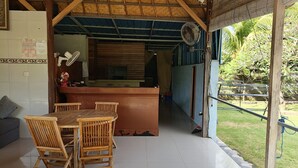 Uma área de refeições ao ar livre coberta, com uma mesa e cadeiras de madeira, um ventilador e vista para um jardim.