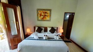 Uma cama bem arrumada com almofadas e um cobertor, uma mesa de cabeceira com abajur, um quadro na parede e uma porta à esquerda.