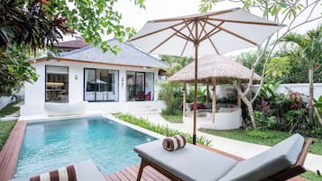 Uma área à beira da piscina com espreguiçadeiras, um guarda-sol de palha e uma casa com janelas amplas e telhado telhado.