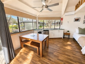 Quarto compacto com piso de madeira, mesa de jantar, cama, cozinha compacta e grandes janelas com vista para o exterior.