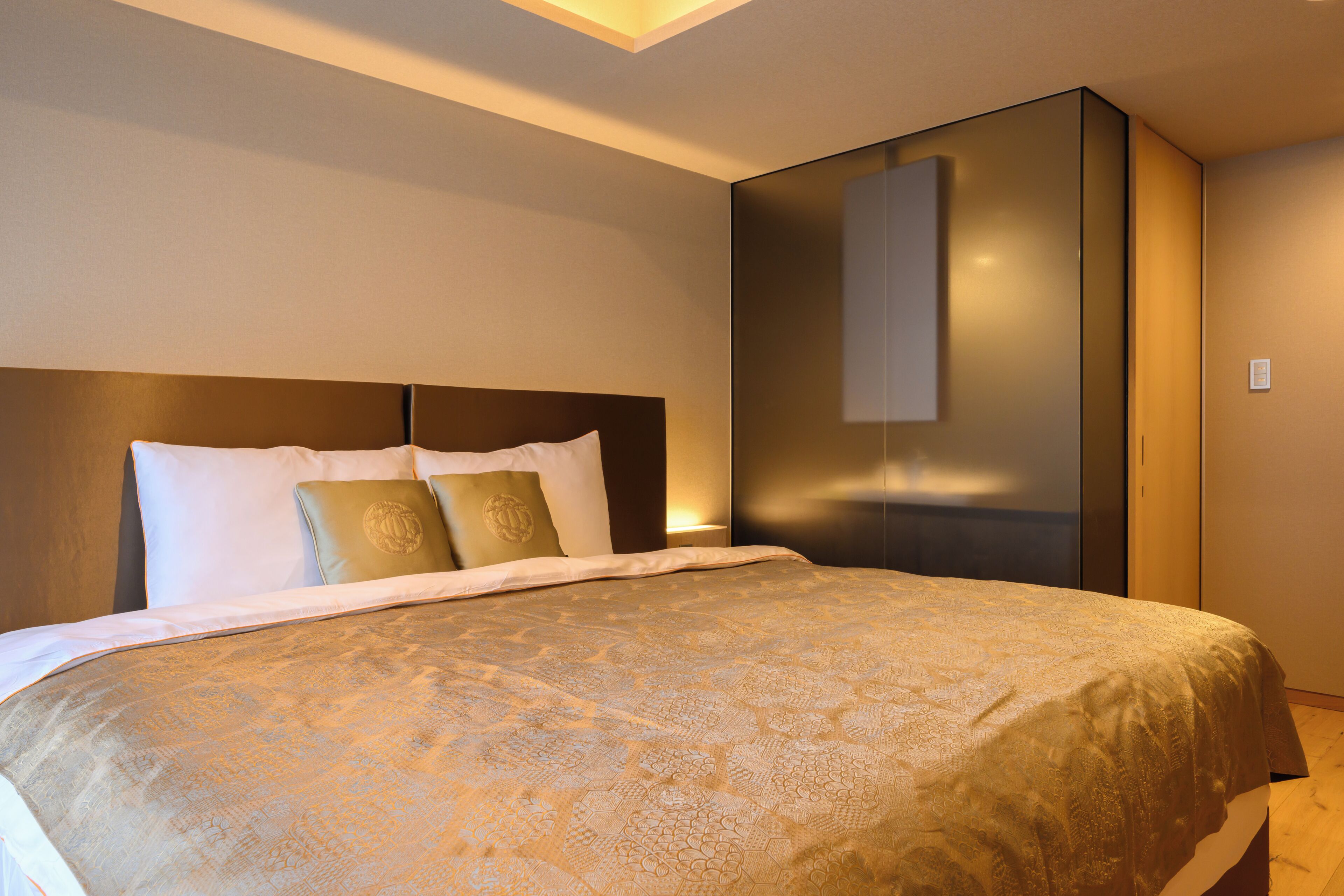 Quarto de hotel moderno com uma cama grande, cabeceira escura e luminária fixada na parede.