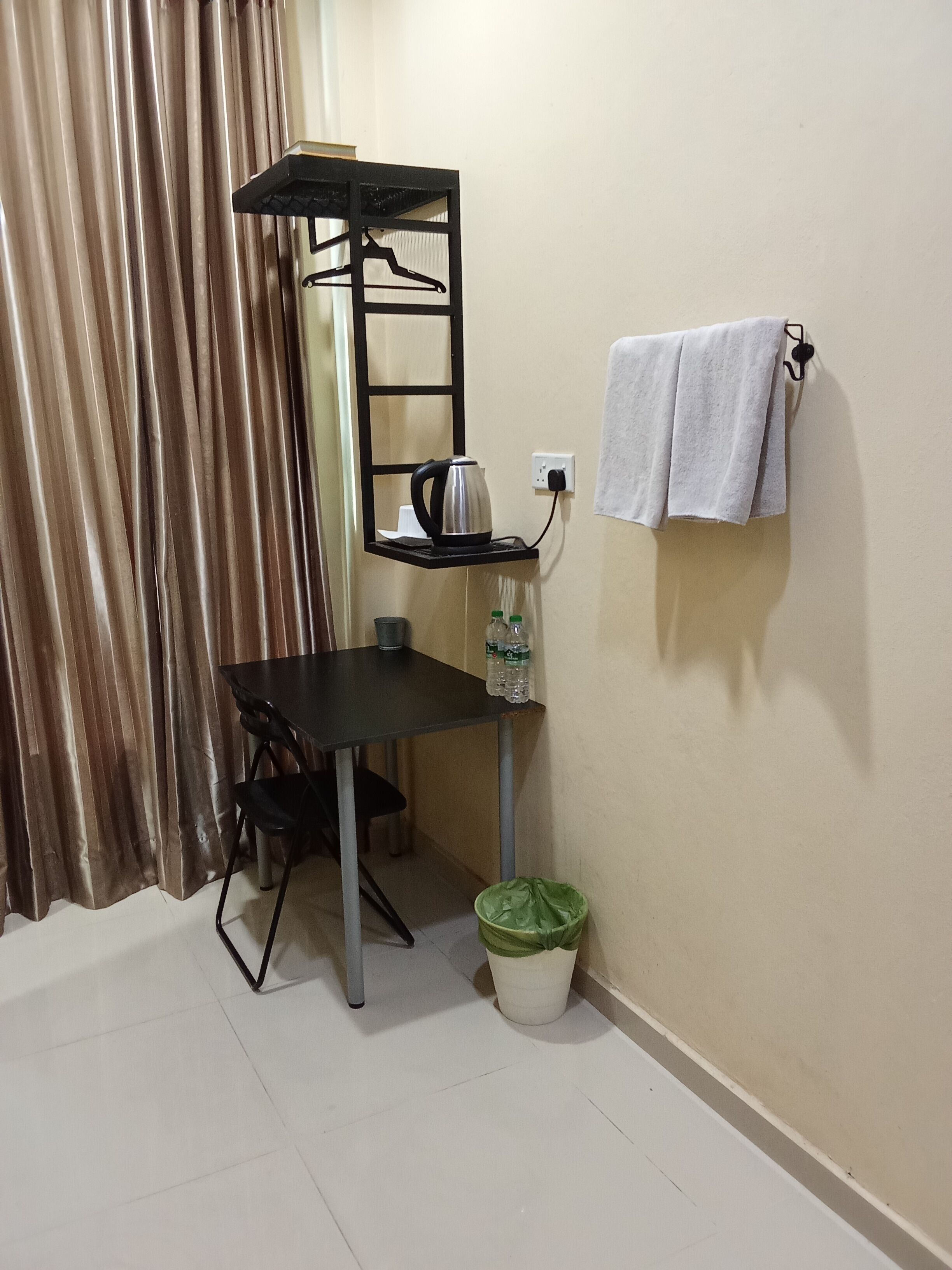 Quarto pequeno com mesa, cadeira, cabide, chaleira, toalha e lixeira.