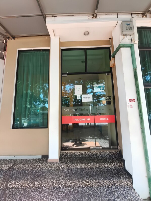 Entrada de um hotel com porta de vidro, uma placa vermelha indicando que é um hotel e uma cortina verde à esquerda.