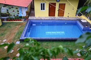 Uma piscina retangular com borda azulejada, cercada por uma área gramada com plantas em vasos.
