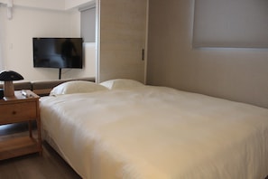 Quarto de hotel com cama, mesa de cabeceira com luminária, televisão e janela com persianas.