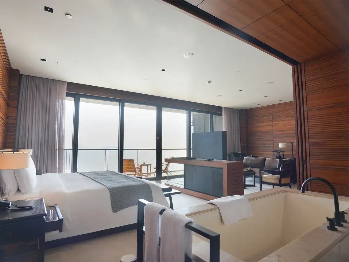 Quarto de hotel moderno com uma cama grande, área de estar com televisão, escrivaninha e uma banheira espaçosa.