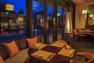 Uma sala de jantar com uma mesa posta para dois, um sofá com almofadas, e vista para a paisagem urbana através de grandes janelas.