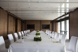 Uma sala de conferência com uma longa mesa, cadeiras brancas e um arranjo floral.