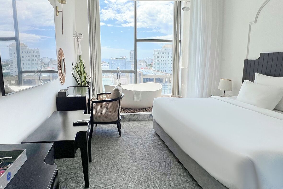 Quarto de hotel com cama, escrivaninha, cadeira, mesa redonda e vista da cidade por grandes janelas.