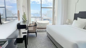 Quarto de hotel com cama, escrivaninha, cadeira, mesa redonda e vista da cidade por grandes janelas.