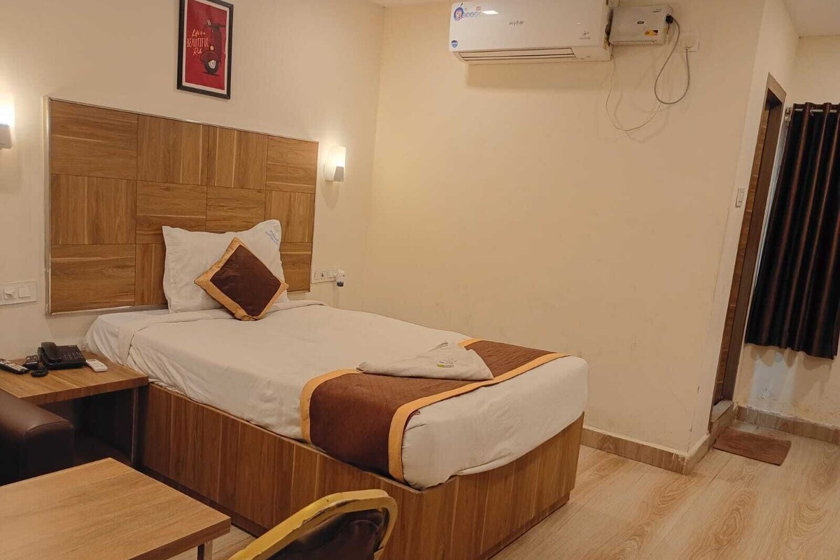 Quarto de hotel com cabeceira de madeira, cama com roupa de cama branca e almofada marrom decorativa, mesa de cabeceira com telefone, uma cadeira e unidade de ar condicionado fixada na parede.