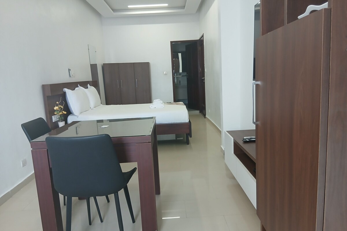 Quarto de hotel com cama, escrivaninha e cadeira, guarda-roupa e banheiro visível através de uma porta aberta.