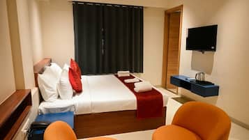 Quarto de hotel com cama, escrivaninha, televisão e duas cadeiras laranja.