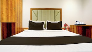 Quarto de hotel com uma cama grande, duas almofadas, cabeceira de madeira, mesa de cabeceira com abajur e uma pequena planta.