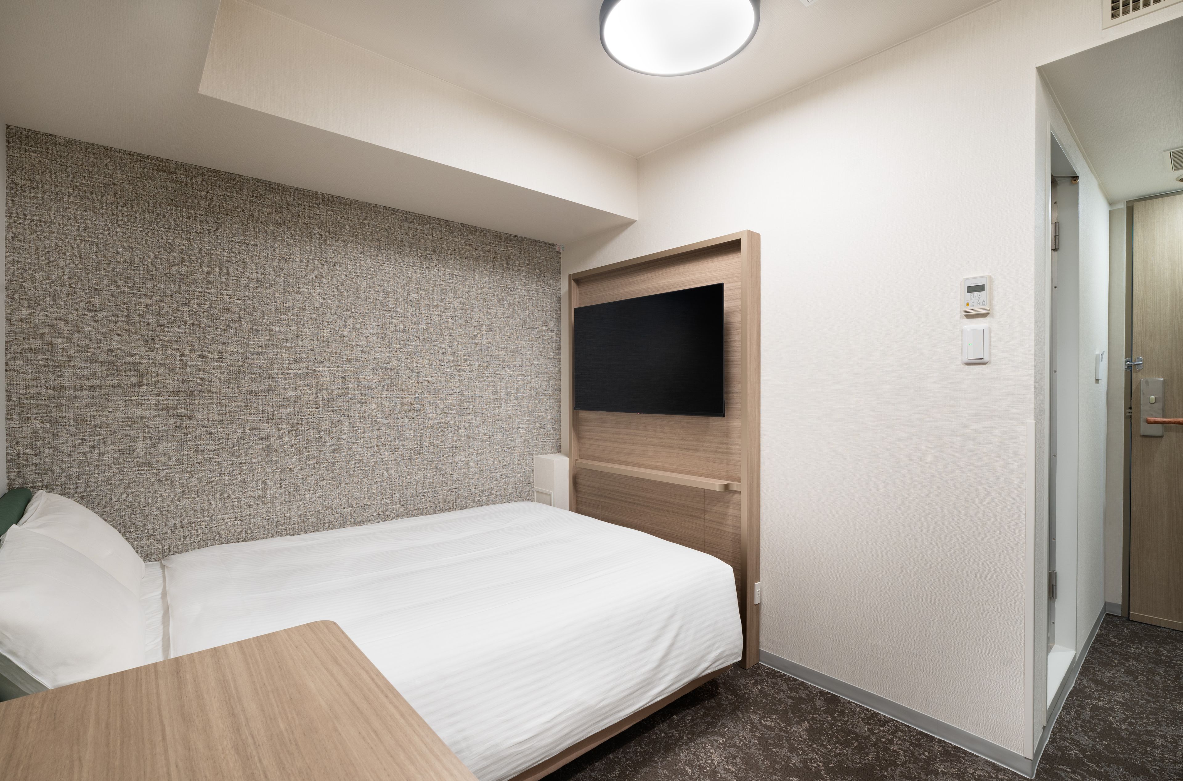 Quarto de hotel com cama, televisão de tela plana na parede, porta e interruptor de luz.