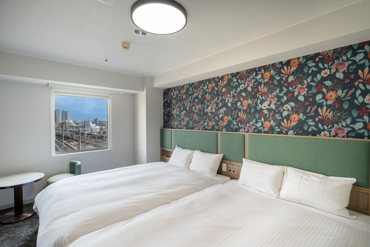 Quarto de hotel com uma cama grande, uma mesa redonda pequena, vista para a cidade através de uma janela e papel de parede floral.