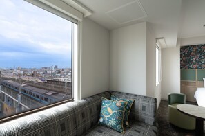 Quarto de canto com uma janela ampla, sofá com almofadas estampadas, mesa pequena e cadeira verde.