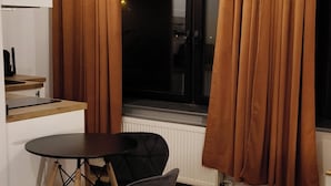 Quarto pequeno com mesa e cadeiras, janela com cortinas, radiador e cama com cobertura branca.