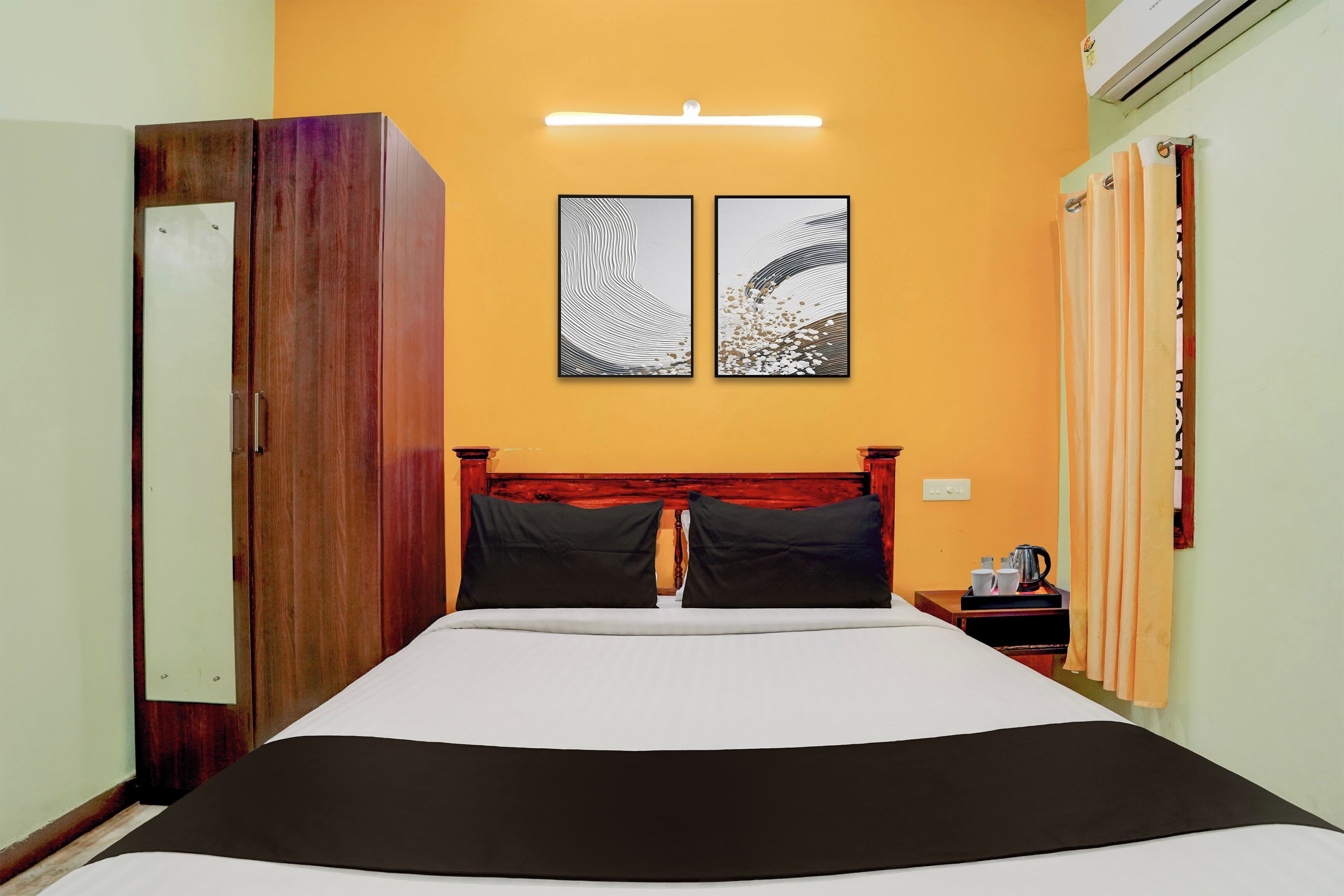 Quarto de hotel com uma cama, duas almofadas, mesa de cabeceira com uma cafeteira e duas obras de arte na parede.