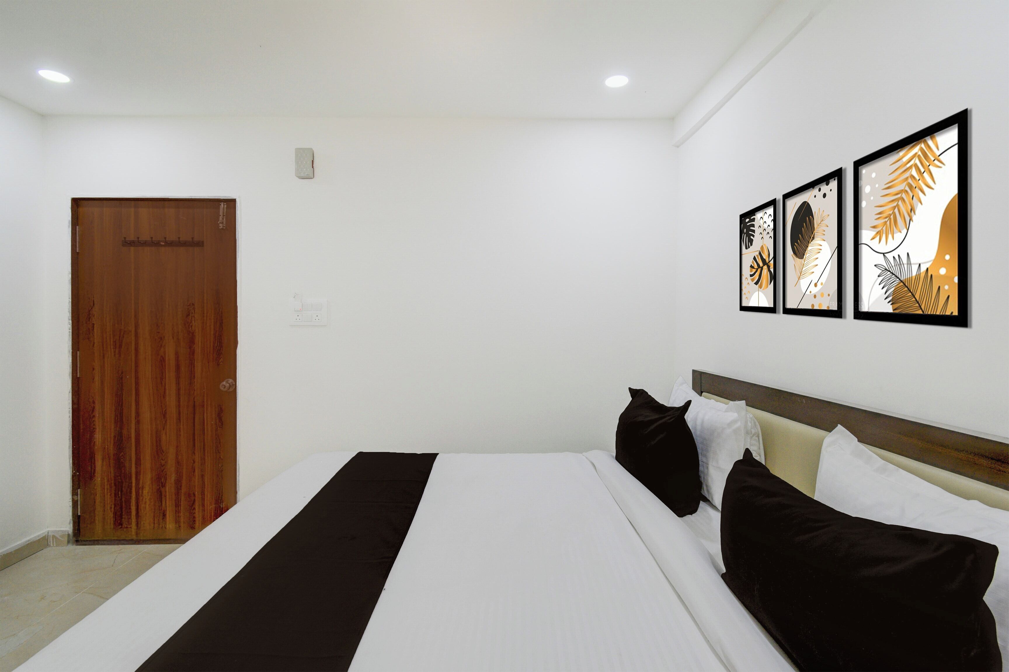 Quarto de hotel com porta de madeira, cama com roupa de cama branca e preta, e três quadros decorativos na parede.