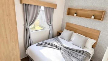 Quarto com cabeceira de madeira, cama com roupa de cama branca, cortinas cinzas e uma pequena prateleira sobre a cama.