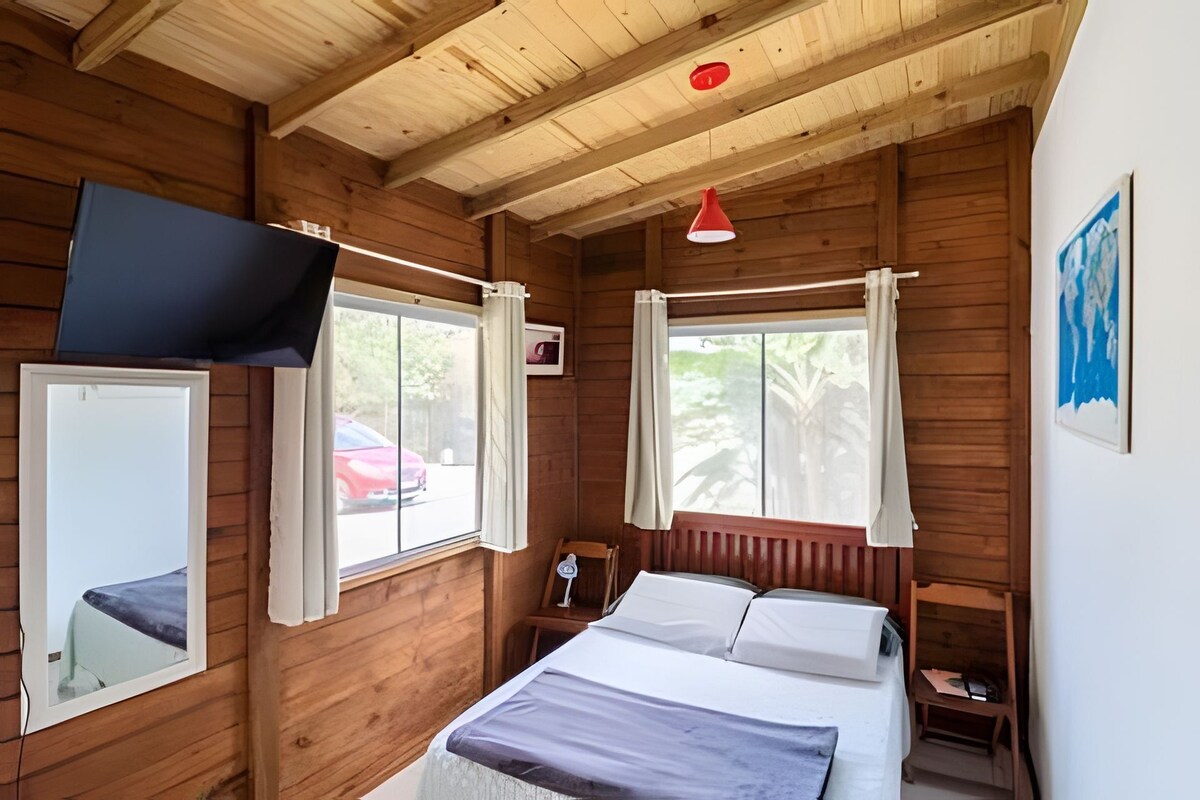 Quarto de cabana de madeira com uma cama, duas janelas, uma luminária de teto vermelha e um quadro pendurado na parede.