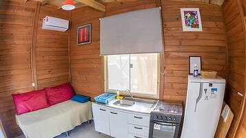 Um quarto compacto com cama, uma kitchenette pequena, janela com persiana e teto de madeira.