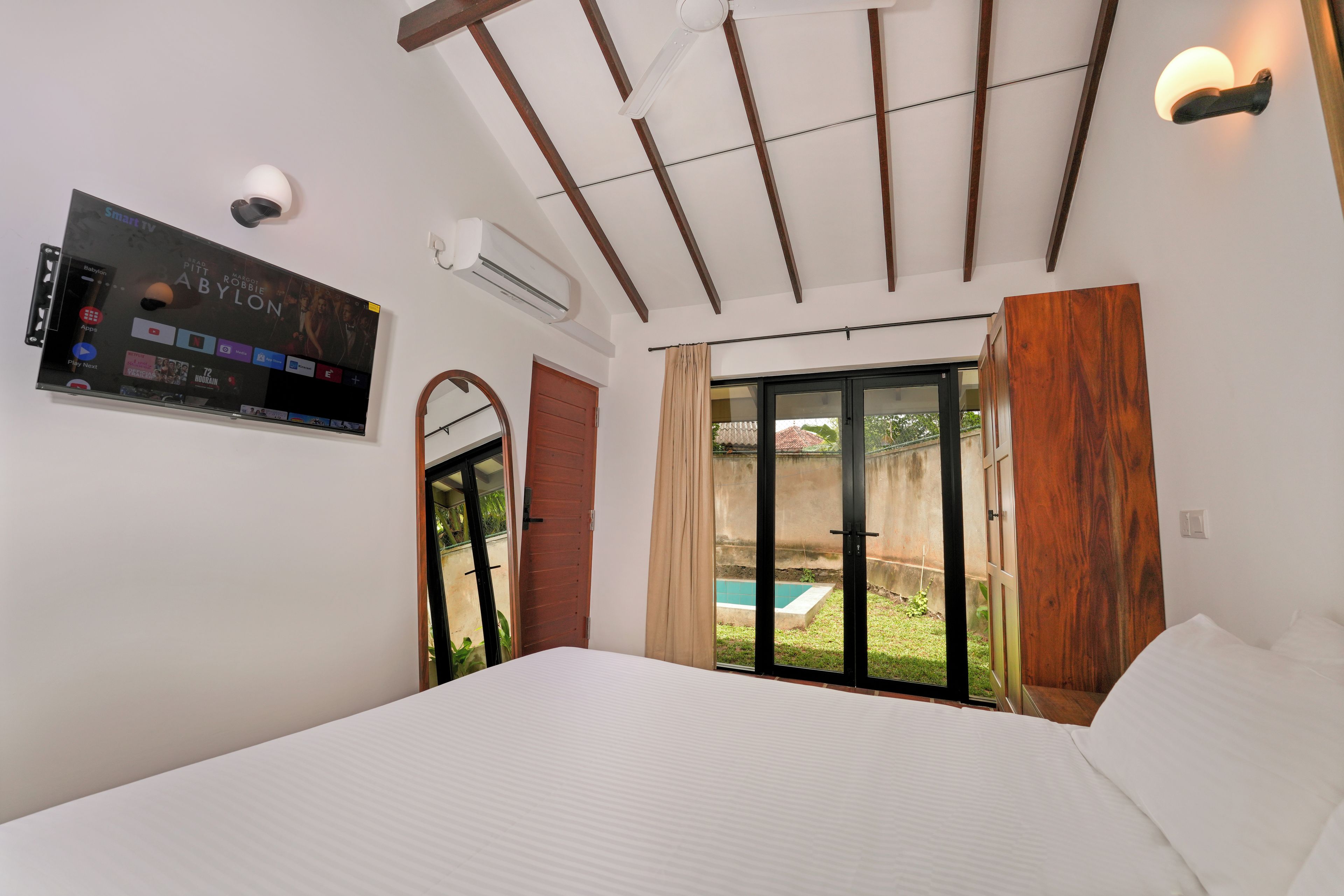 Quarto com cama, TV na parede, aparelho de ar condicionado e vista para uma piscina através de uma porta de vidro.
