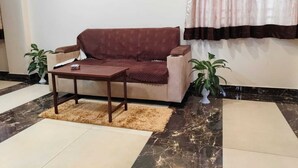 Uma sala de estar com sofá, mesa de centro, duas plantas em vasos e uma janela com cortinas.