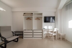 Um quarto com um armário branco, televisão na parede, uma escrivaninha com cadeiras e um sofá.