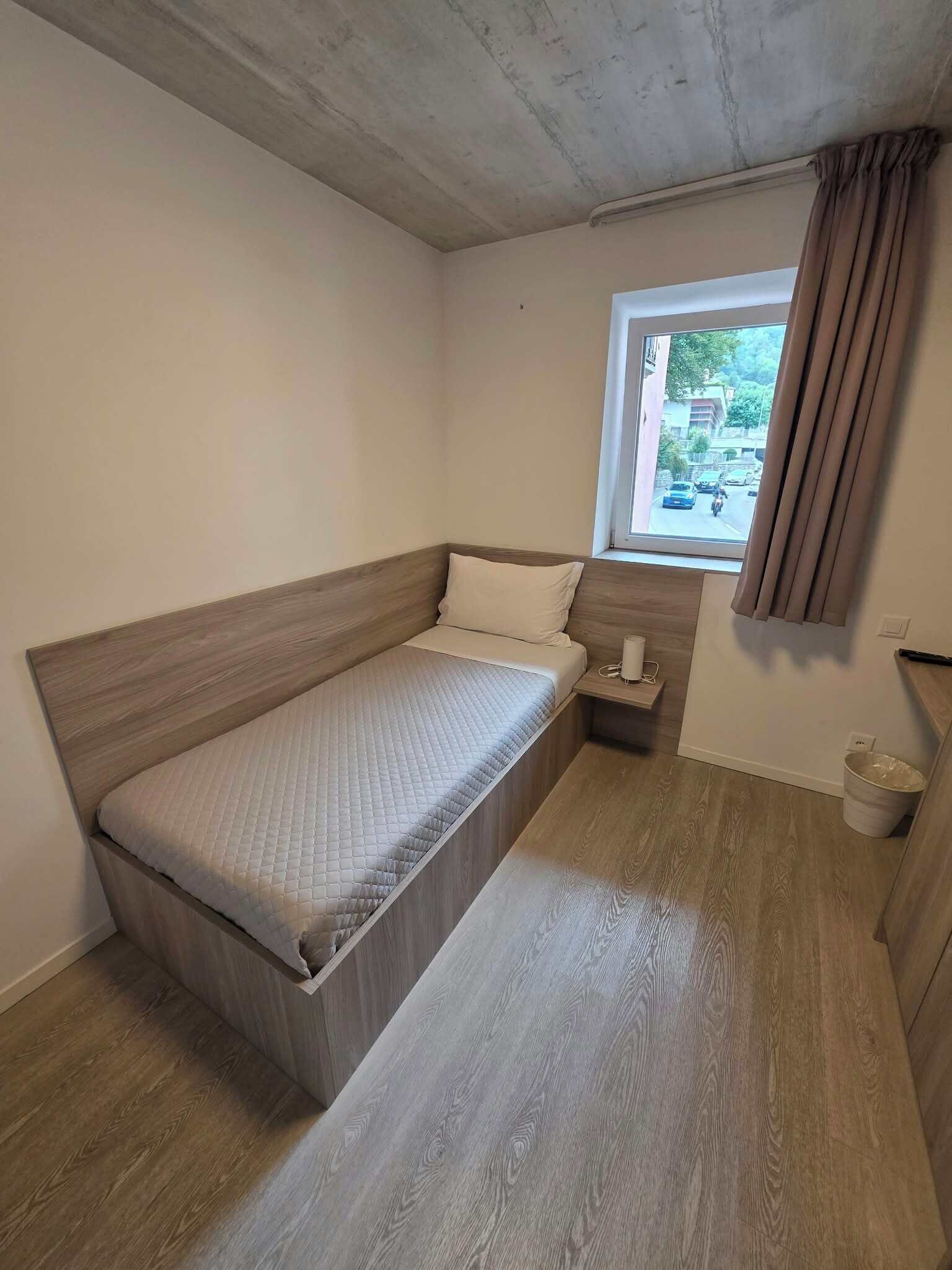 Quarto pequeno com cama de solteiro, cabeceira de madeira, mesa de cabeceira com luminária e janela com cortinas.