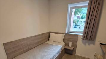 Quarto pequeno com cama de solteiro, cabeceira de madeira, mesa de cabeceira com luminária e janela com cortinas.
