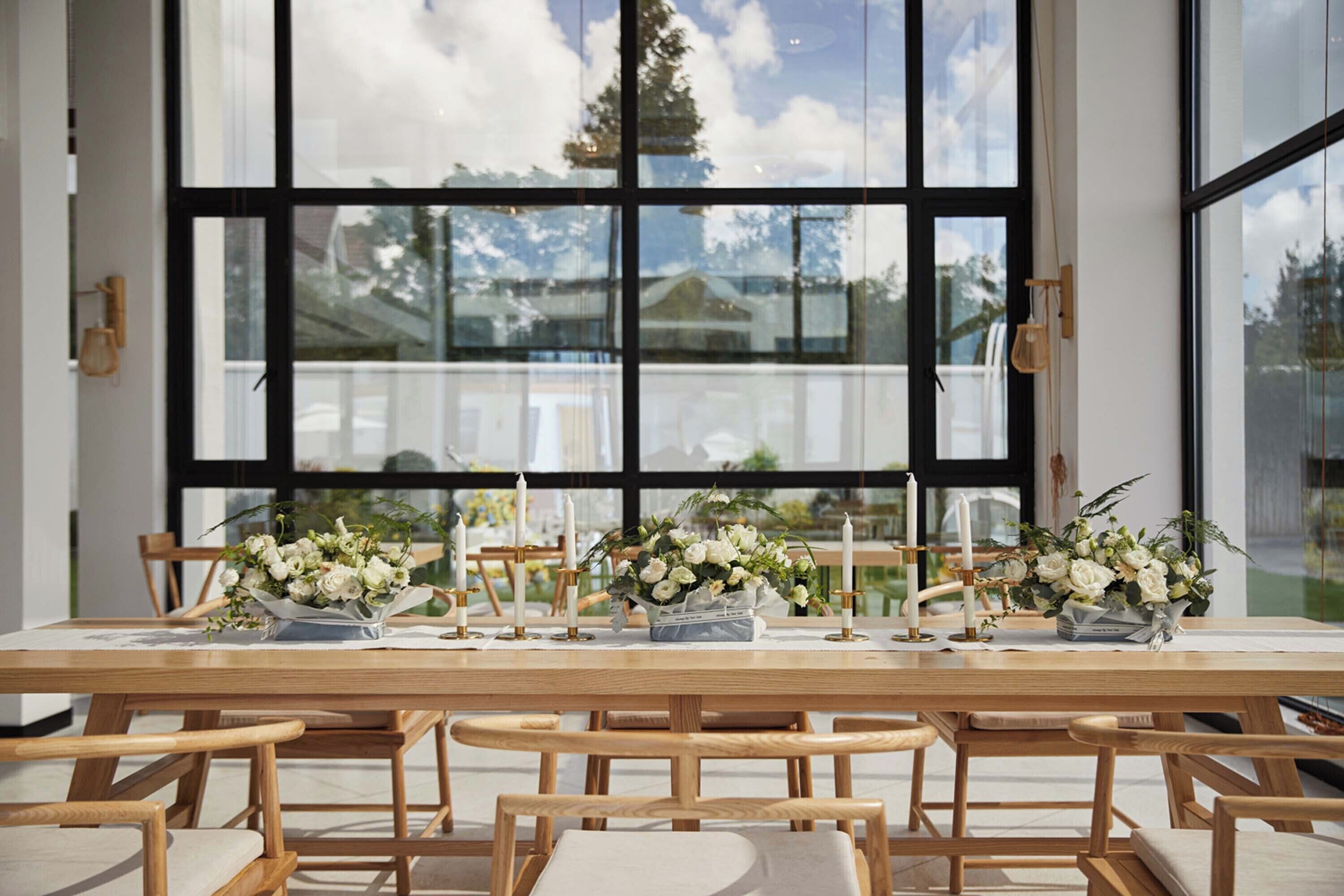 Mesa de jantar posta com cadeiras, velas e arranjos florais em um quarto com grandes janelas.
