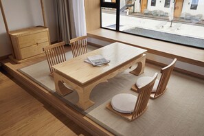 Uma mesa de jantar de madeira com quatro cadeiras, um pequeno armário de madeira e vista para o exterior através de uma grande janela.