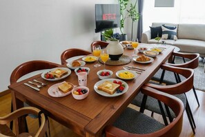 Uma mesa de jantar posta com itens de café da manhã, uma televisão exibindo 'NETFLIX' e um sofá ao fundo.